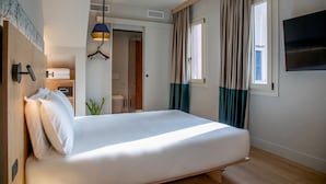 Quarto de hotel com uma cama, um guarda-roupa, uma luminária pendente, uma janela com cortinas e uma televisão na parede.