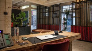 Uma mesa de recepção com um laptop, telefone e um copo de água. Há plantas e um divisória vermelha ao fundo.