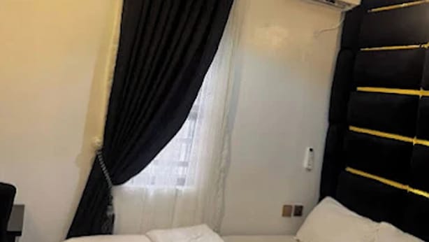 Quarto com cama, janela com cortinas pretas, aparelho de ar condicionado e cabeceira preta com detalhes amarelos.