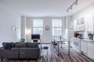 Um apartamento moderno com um sofá cinzento, uma televisão, uma pequena mesa de jantar com banquetas e uma cozinha com armários e eletrodomésticos brancos.