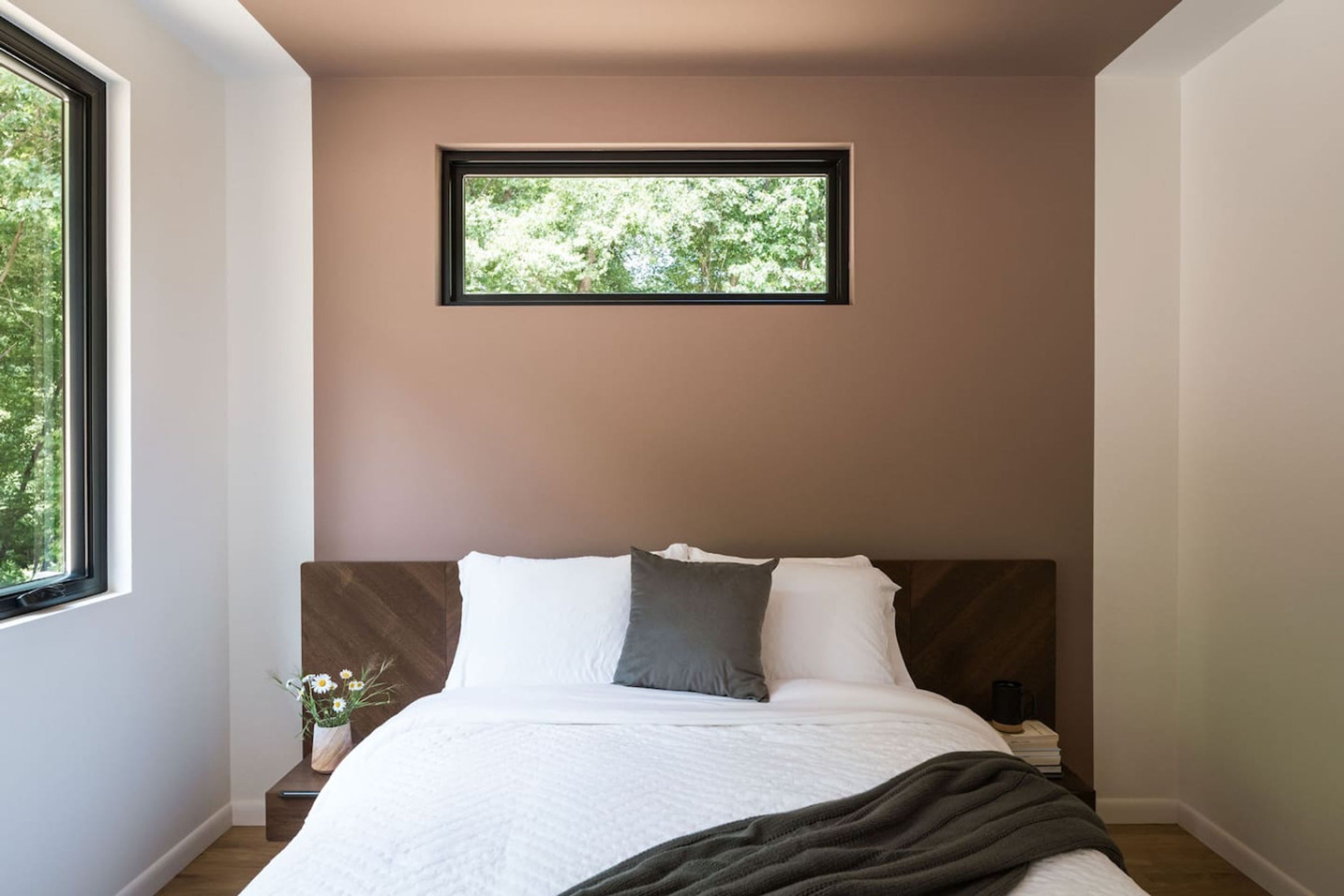 Quarto com uma janela grande, cama com roupa de cama branca e cinza, mesa de cabeceira com abajur e vista para árvores lá fora.