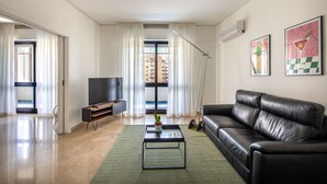 Uma sala de estar moderna com um sofá de couro preto, uma televisão de tela plana, uma mesa de centro e uma janela grande com cortinas.
