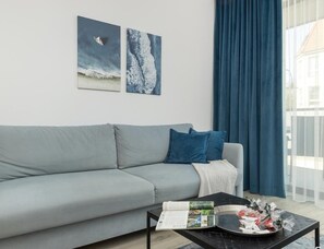 Uma sala de estar moderna com um sofá cinza, duas almofadas azuis e uma mesa de centro com revistas e um prato de comida.