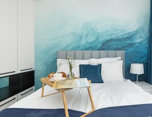 Quarto de hotel com cama, mesa de cabeceira com bandeja de itens de café da manhã, televisão e um mural na parede.