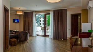 Quarto de hotel com piso de madeira, área de refeições com mesa e cadeiras, sofá, televisão e varanda com cortinas.