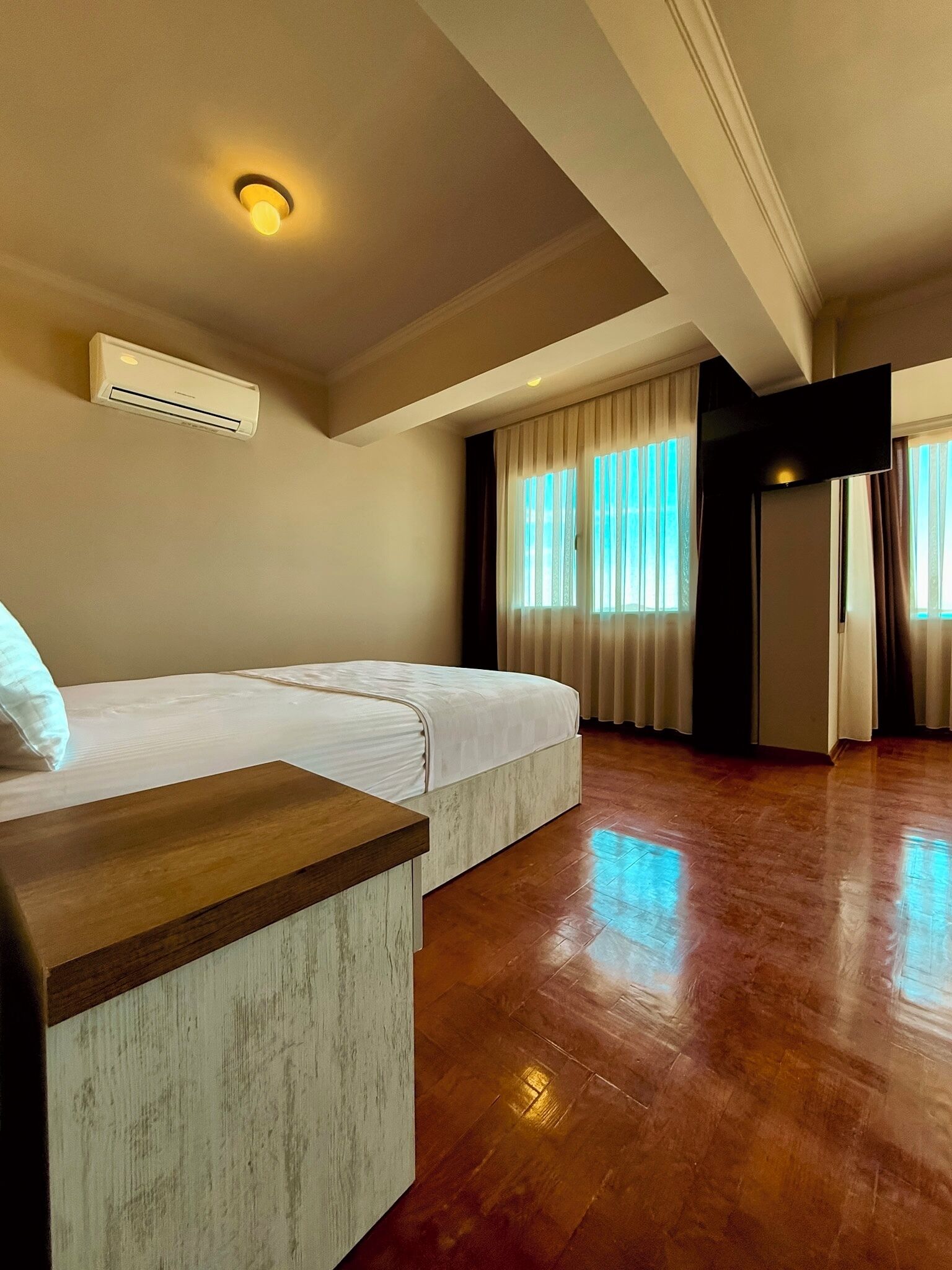 Quarto de hotel com cama de madeira, mesa de cabeceira, ar condicionado no teto e grandes janelas com cortinas.