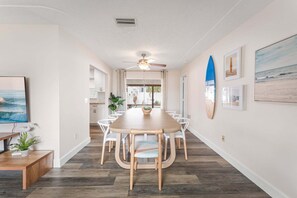 Uma sala de jantar com uma mesa comprida, cadeiras brancas e um ventilador de teto.