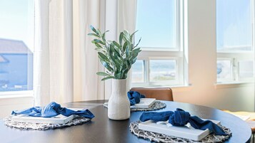 Mesa de jantar com um vaso branco texturizado contendo folhas verdes, guardanapos azuis e um objeto retangular branco sobre um sousplat trançado.