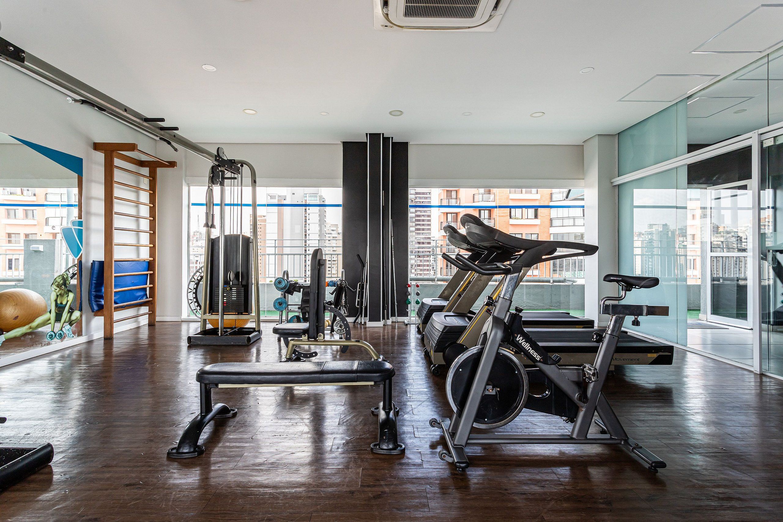 Academia com equipamentos de ginástica, um banco e vista para edifícios através de amplas janelas.
