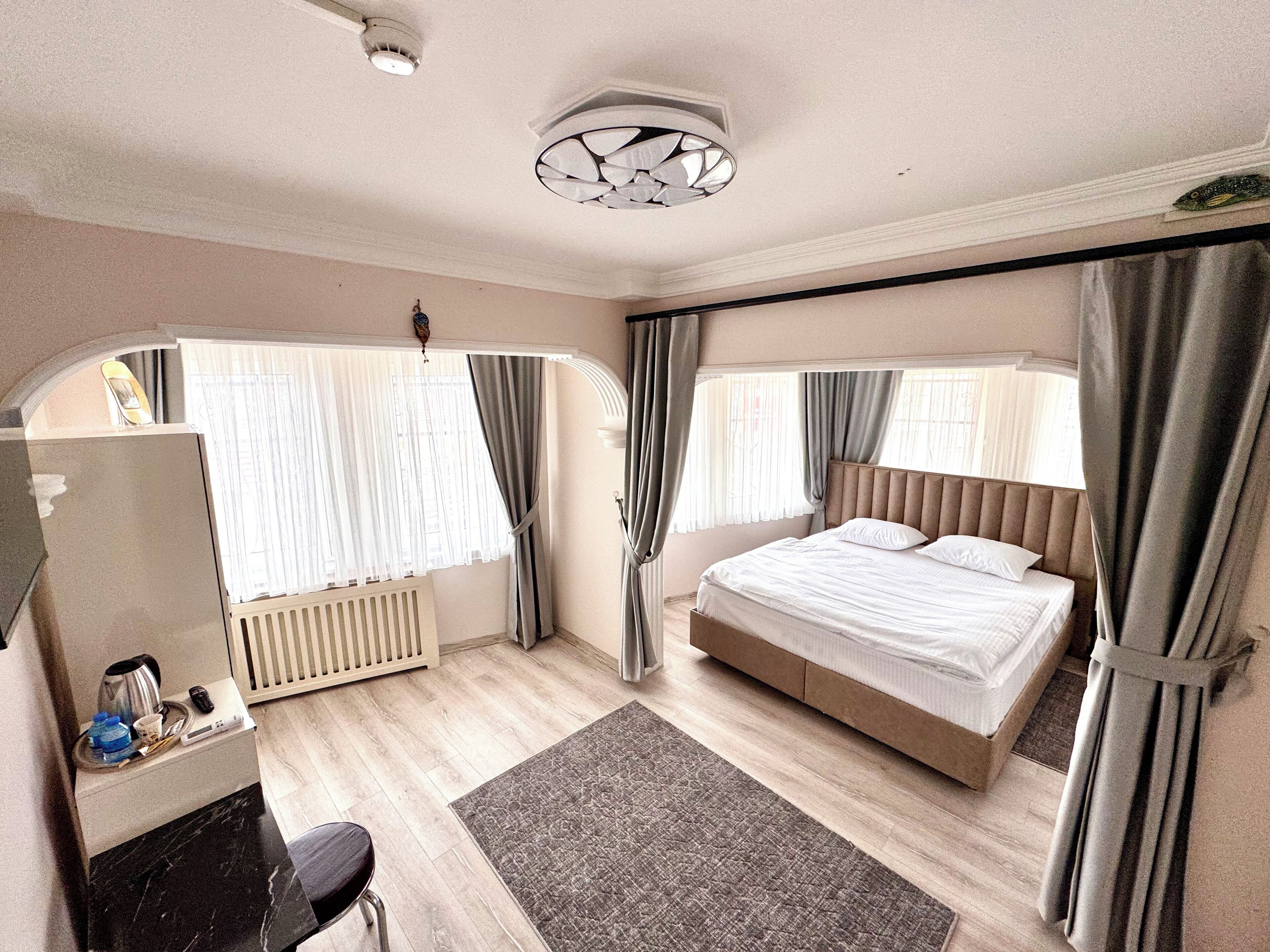 Quarto de hotel com cama, escrivaninha, cadeira, radiador e janela com cortinas.