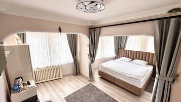 Quarto de hotel com cama, escrivaninha, cadeira, radiador e janela com cortinas.