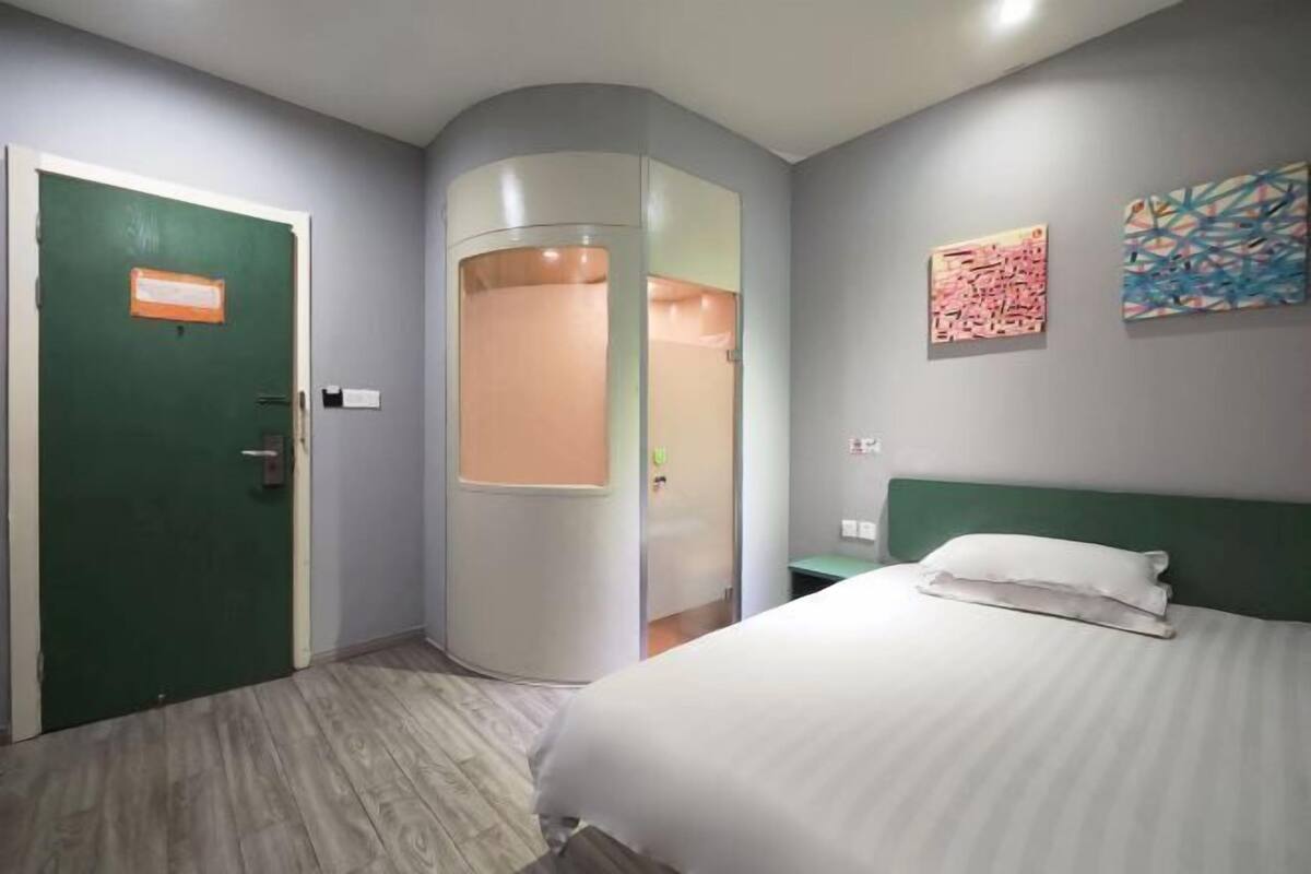 Quarto de hotel com cama, porta verde, espelho curvo e duas pinturas abstratas na parede.