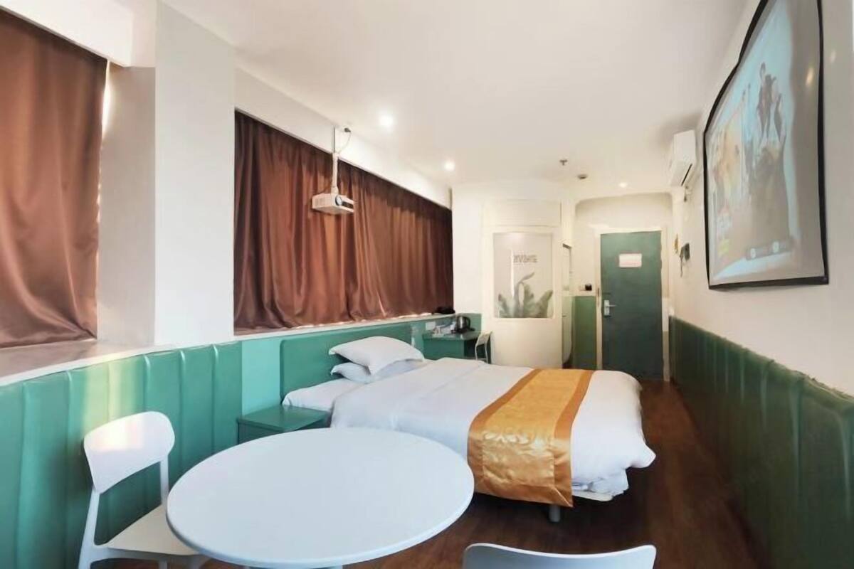 Quarto de hotel com cama, mesa redonda, cadeira, janela com cortinas, porta e projetor fixado na parede.