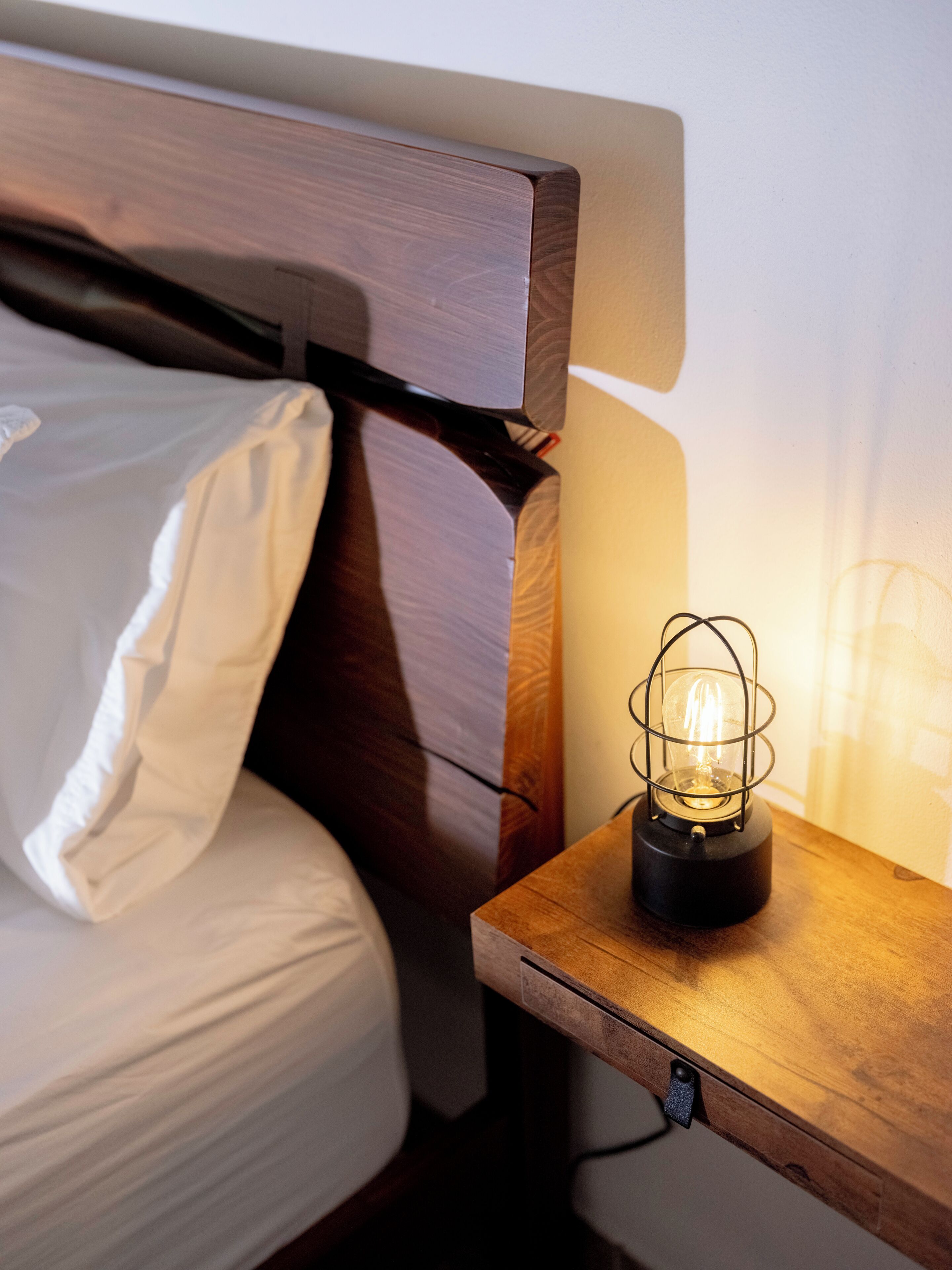 Uma cama com cabeceira de madeira, mesa de cabeceira com abajur e travesseiro branco.