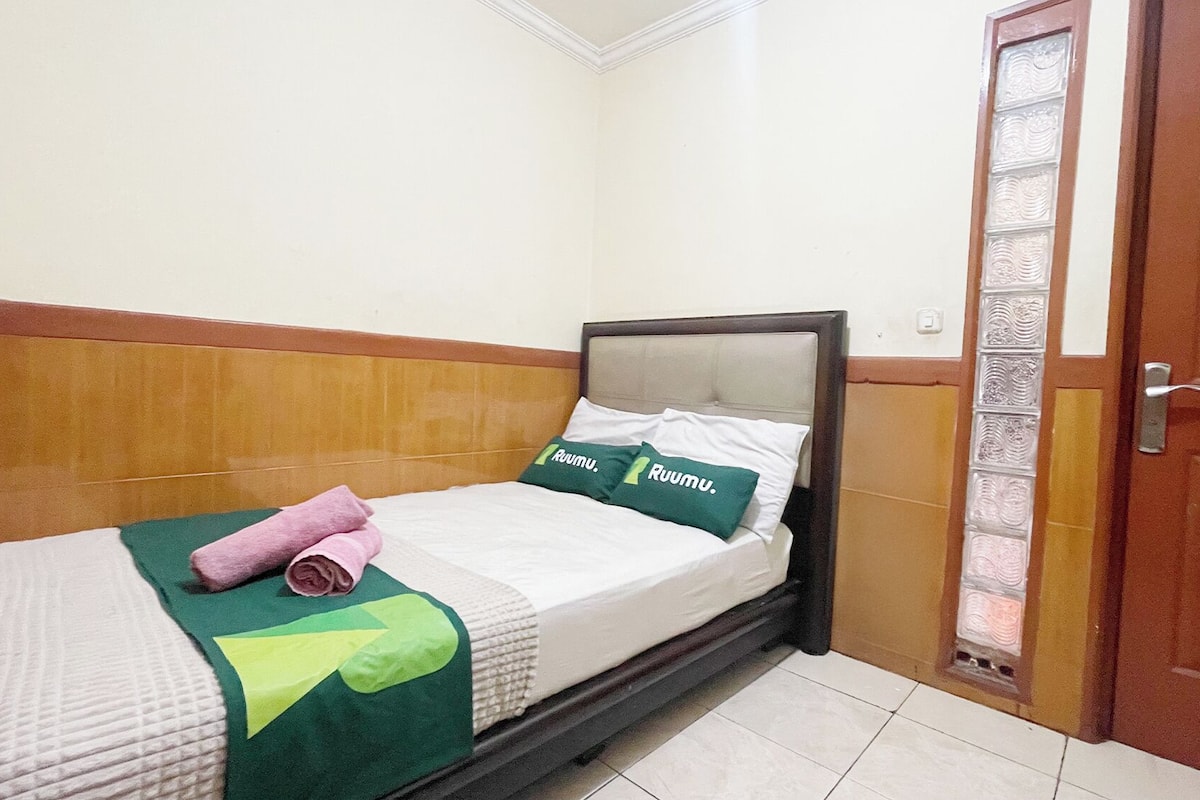 Uma cama de solteiro com cabeceira, um cobertor verde e amarelo, duas almofadas com a marca 'Ruumu', uma toalha rosa e uma porta de madeira com painel de vidro.