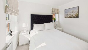 Quarto com uma cama grande, mesas de cabeceira brancas, cabeceira preta, janela com cortinas e um quadro na parede.