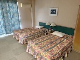 Quarto de hotel com duas camas, cada uma com roupa de cama florida, mesa de cabeceira com luminárias, ar condicionado fixado na parede e um quadro emoldurado.