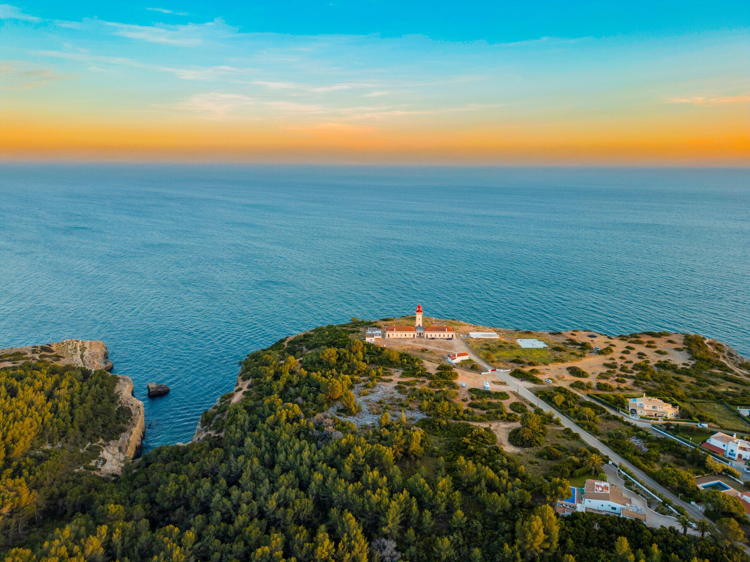 Vista aérea de uma área costeira com um farol, um pequeno povoado e uma visão clara do mar.