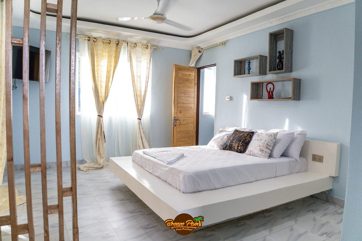 Quarto com cama, cortinas, porta de madeira e estantes com objetos decorativos.