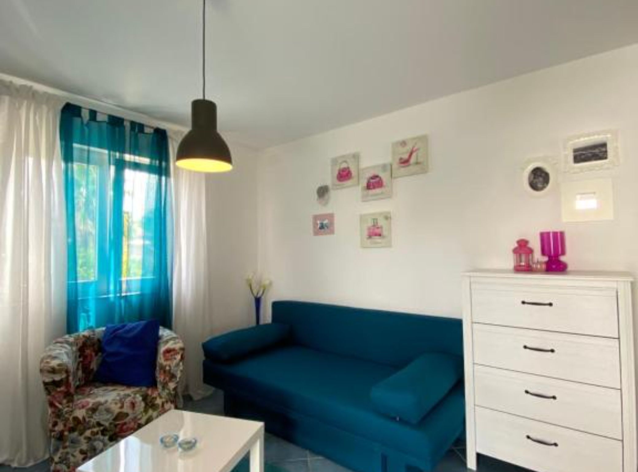 Um quarto com um sofá azul, uma poltrona estampada, uma cômoda branca e um lustre.