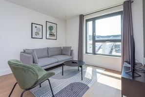 Uma sala de estar moderna com um sofá cinzento, uma poltrona verde, uma mesa redonda pequena, uma televisão de tela plana e uma grande janela com cortinas.