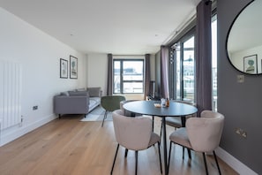 Uma sala de estar moderna com um sofá cinzento, uma mesa redonda com cadeiras, uma janela grande com cortinas e um espelho na parede.