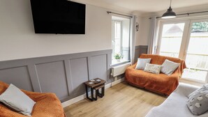 Quarto com dois sofás, um laranja e outro cinza, uma mesinha e uma TV fixada na parede.