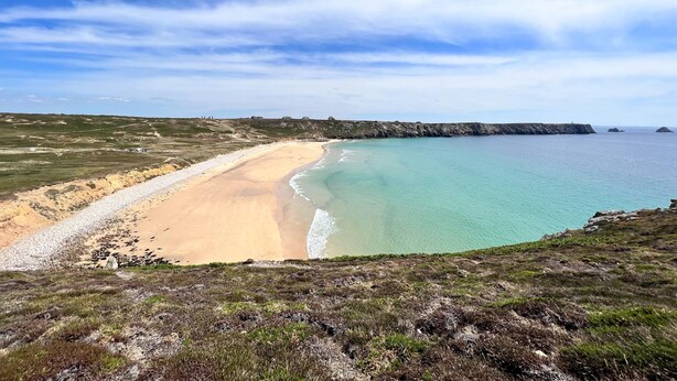 Paisagem costeira com uma praia de areia, águas turquesa cristalinas e uma colina gramada em primeiro plano.