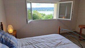 Um quarto com cama, abajur, janela com vista aberta para um lago e árvores, e uma mesa pequena.