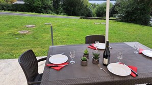 Mesa de jantar ao ar livre montada com pratos, talheres e copos de vinho, rodeada por cadeiras, com vista para uma área gramada e árvores ao fundo.