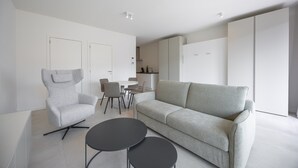 Uma sala de estar moderna com um sofá cinzento, uma poltrona cinzenta e duas mesas de centro pretas. Há uma área de jantar com uma mesa redonda e cadeiras. O quarto tem paredes brancas e um guarda-roupa grande.