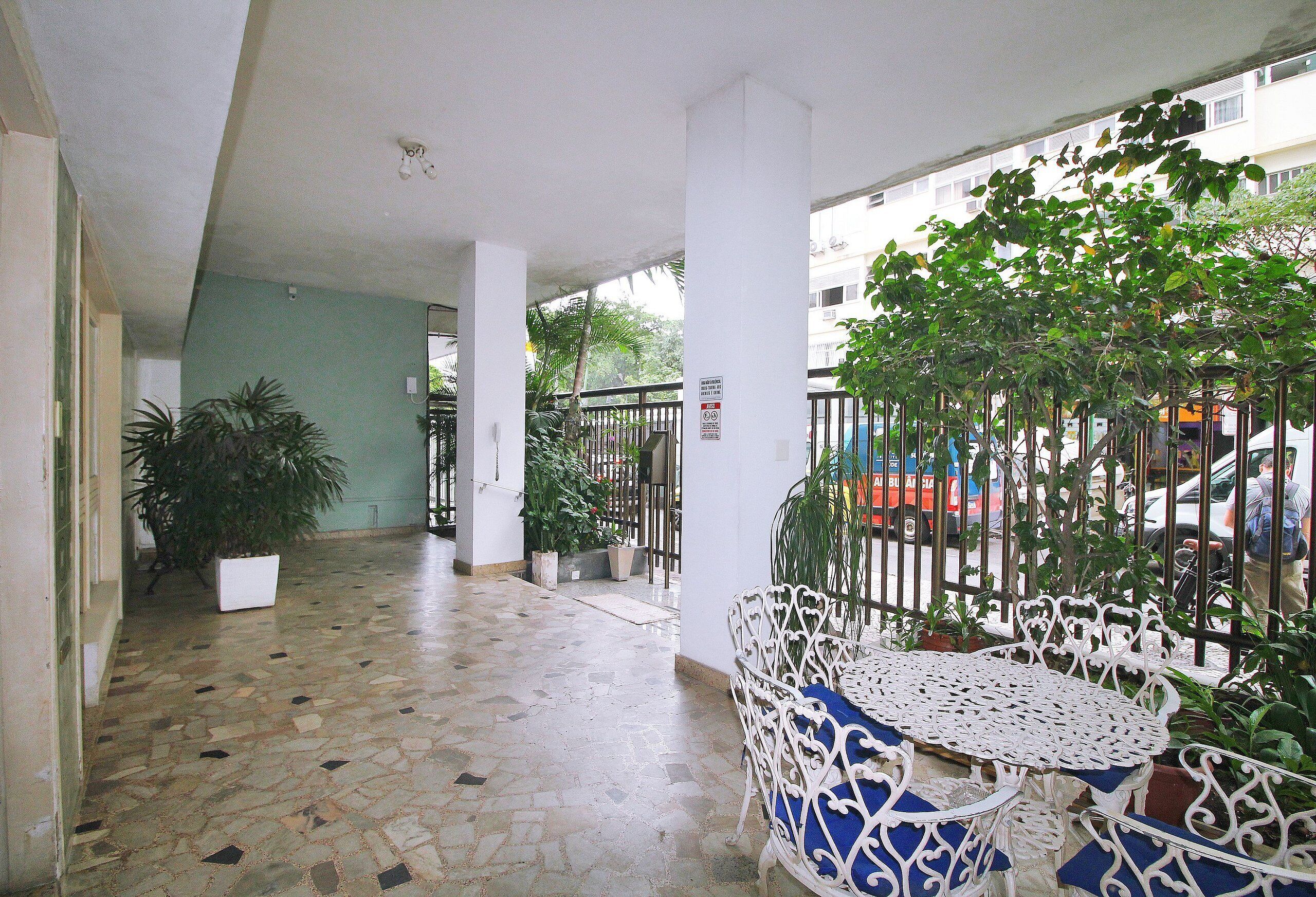 Corredor com piso de azulejos, pilares brancos e guarda-corpo de metal, contendo uma mesa e cadeiras.