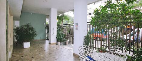 Corredor com piso de azulejos, pilares brancos e guarda-corpo de metal, contendo uma mesa e cadeiras.