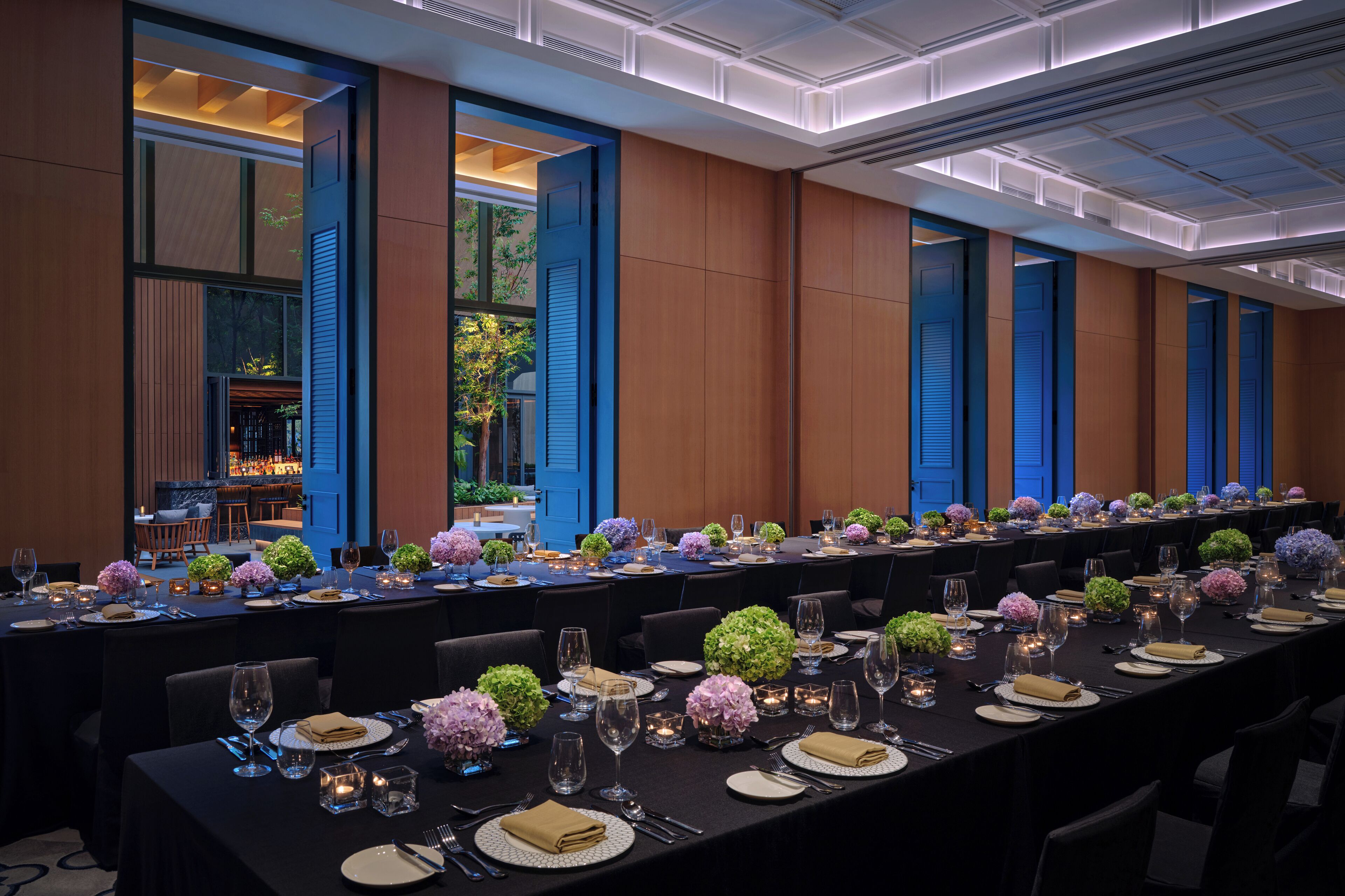 Uma longa mesa de jantar posta para um evento formal, com diversas cadeiras, velas e arranjos florais.