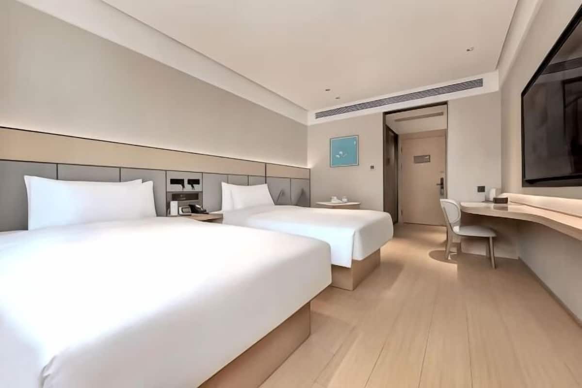 Quarto de hotel com duas camas, uma mesa, uma cadeira, televisão e porta que dá para outro espaço.