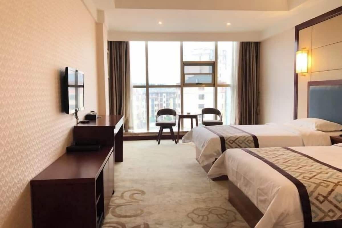 Quarto de hotel com duas camas, uma escrivaninha, uma cadeira, uma televisão e uma janela ampla com vista para edifícios.
