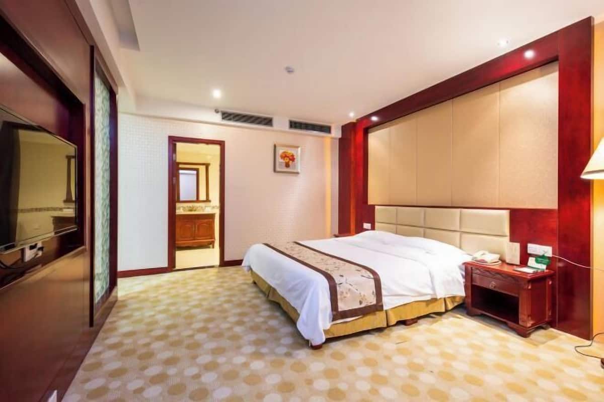 Quarto de hotel com uma cama grande, televisão, mesa de cabeceira com abajur e banheiro visível através de uma porta aberta.