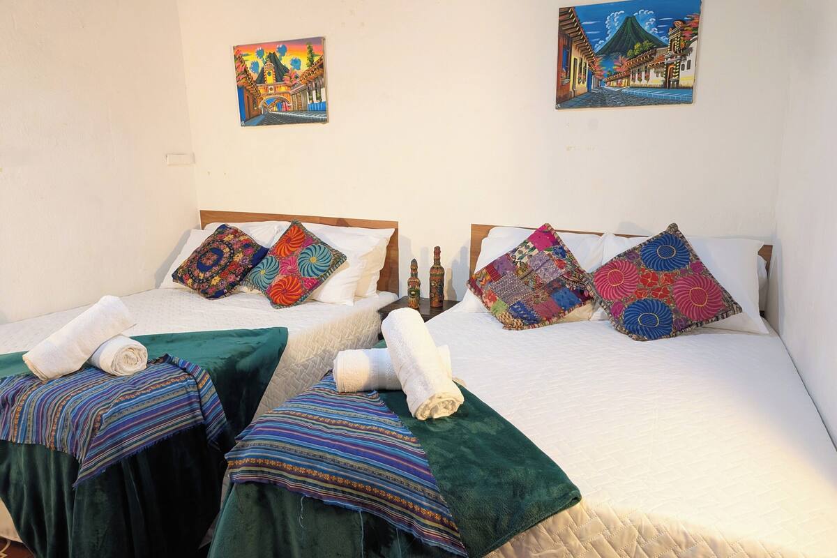 Quarto de hotel com duas camas, cada uma com almofadas coloridas e um cobertor verde. Há duas pinturas na parede acima das camas.