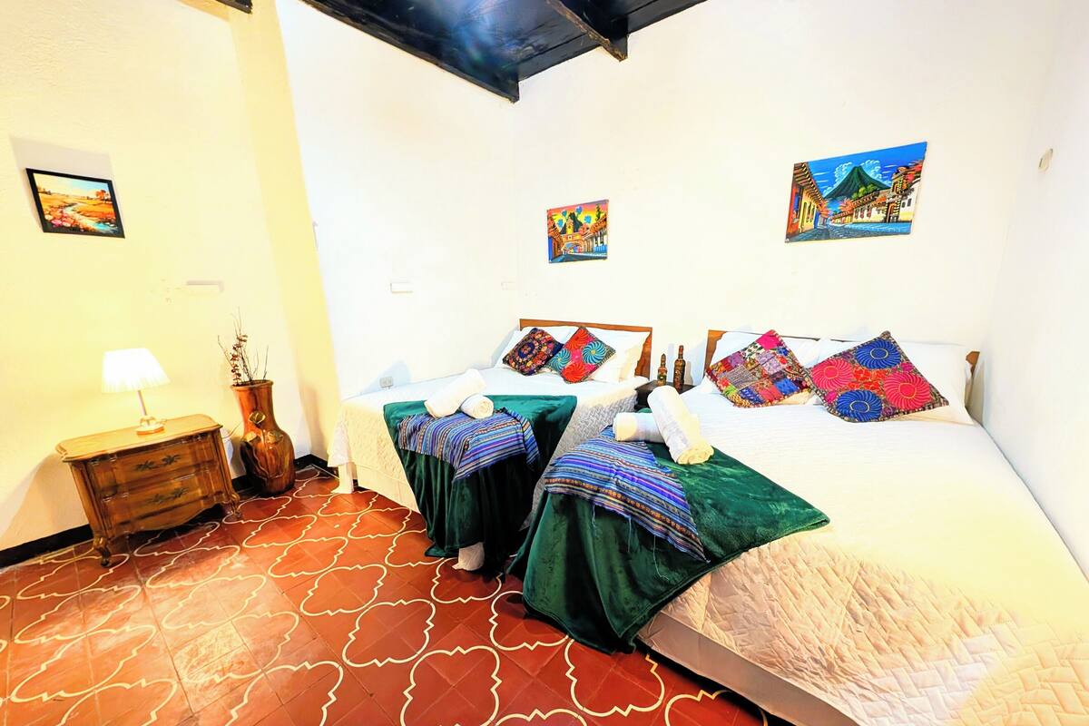 Quarto com duas camas, cada uma com colcha verde e várias almofadas coloridas. Há uma mesa de cabeceira de madeira com abajur e um vaso com plantas secas. O chão apresenta um design vermelho estampado.