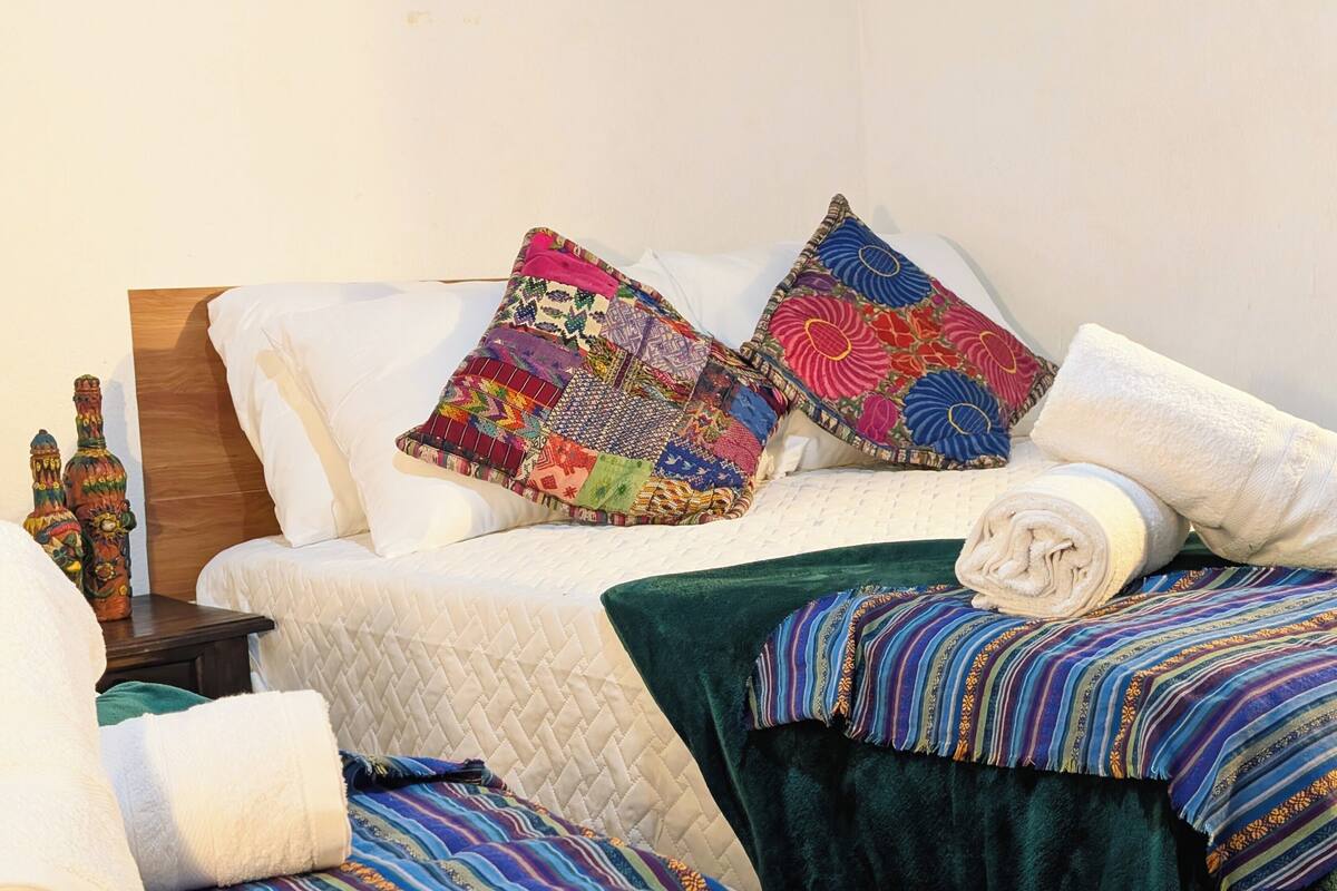 Uma cama com edredom branco acolchoado, duas almofadas coloridas, um cobertor verde e duas toalhas brancas dobradas.