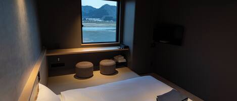 Quarto de hotel moderno com uma janela grande, uma cama com lençóis brancos, dois pufes redondos e uma pequena mesa de cabeceira com uma luminária.