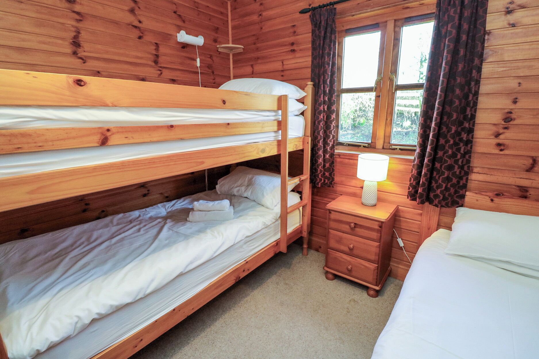 Quarto de cabana de madeira com beliches, mesa de cabeceira com abajur e janela com cortinas.