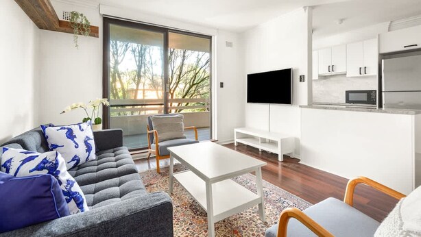 Uma sala de estar moderna com um sofá, mesa de centro e uma televisão. Há uma área de cozinha com armários e um micro-ondas.
