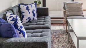 Uma sala de estar moderna com um sofá cinzento, almofadas estampadas em azul, uma mesa branca e vista para árvores através de uma grande janela.
