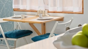 Uma sala de jantar com uma mesa posta para dois, uma janela com cortinas leves e uma tigela com maçãs verdes sobre a mesa.