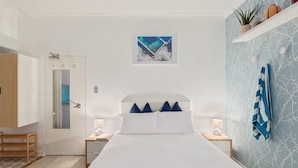 Quarto com cama, duas mesas de cabeceira, um criado-mudo, uma porta, um espelho, uma prateleira na parede com objetos decorativos e uma obra de arte.