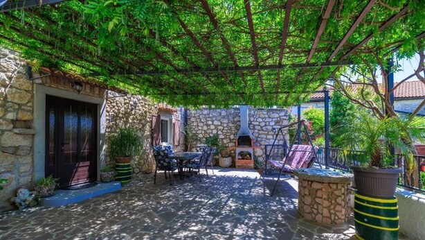 Um pátio com paredes de pedra, pérgola coberta de vegetação, área de estar ao ar livre e lareira de pedra.