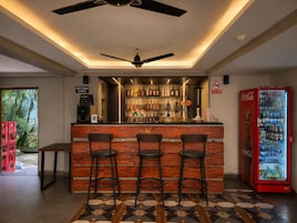 Uma área de bar com balcão de madeira, três banquetas, um ventilador de teto e uma máquina de refrigerantes Coca-Cola.