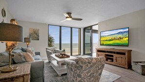 Uma sala de estar moderna com uma televisão grande, assentos confortáveis, um ventilador de teto e vista para a praia.
