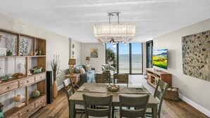 Uma sala de jantar com mesa posta para quatro pessoas, um candelabro e vista para o oceano através de uma grande janela.